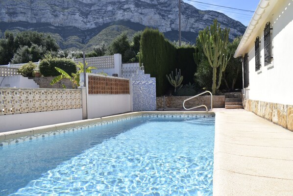 Uma piscina com água azul cristalina, cercada por um edifício branco com borda de azulejos, um muro de pedra e um cacto.