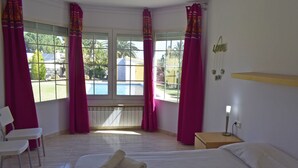 Quarto de hotel com cama, duas cadeiras, mesa de cabeceira com luminária, janela com cortinas e vista para uma piscina.