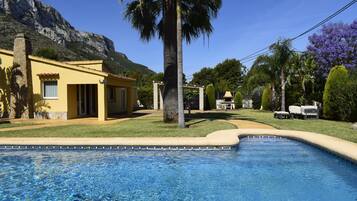Piscina com montanhas ao fundo, um edifício amarelo e vegetação bem cuidada.