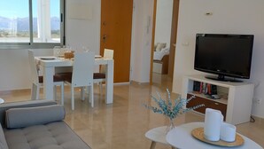 Uma sala de estar moderna com um sofá, uma mesa de jantar com cadeiras, uma televisão em um móvel e vista para montanhas através de uma janela.
