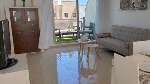 Uma sala de estar moderna com sofá, mesa de jantar e televisão. Há uma varanda com vista para edifícios e um jardim.