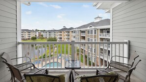 Varanda com duas cadeiras e uma mesa pequena, com vista para uma piscina e uma área ajardinada, com edifícios ao fundo.