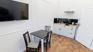 Uma cozinha compacta com piso de madeira em padrão herringbone, armários brancos e uma TV de tela plana fixada na parede.