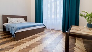 Quarto com piso de madeira em padrão herringbone, uma cama com manta azul, mesa de cabeceira e janela com cortinas verde-água.
