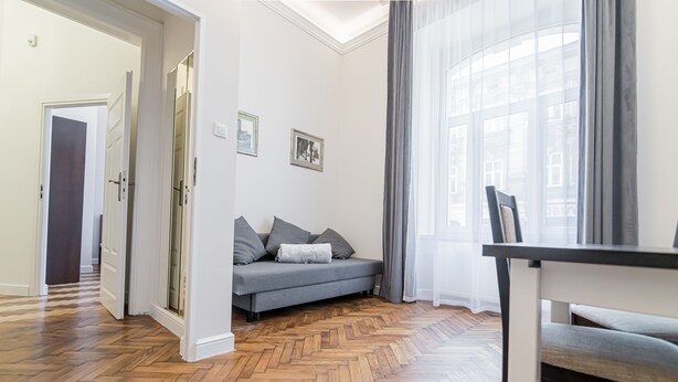 Uma sala de estar moderna com piso de madeira em padrão herringbone, um sofá cinza com almofadas, uma mesa de jantar com cadeiras e janelas amplas com cortinas cinzas.