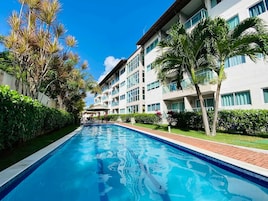Uma piscina com superfície de água azul cristalina, rodeada por vegetação exuberante e um edifício com varandas.