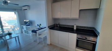 Cozinha moderna com armários brancos, bancada preta e forno embutido. Possui área de jantar com mesa e cadeiras brancas, e uma varanda com vista para a água.