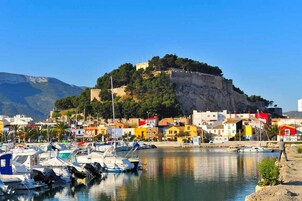 Uma cidade costeira com edifícios coloridos, uma marina com barcos e uma colina imponente com uma fortificação.