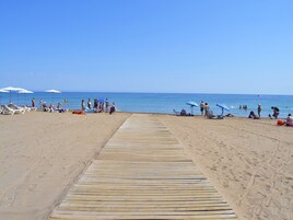 Um calçadão de madeira que leva a uma praia arenosa, com guarda-sóis e espreguiçadeiras.