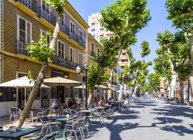 Um café ao ar livre com mesas e cadeiras, cercado por edifícios com varandas e árvores.