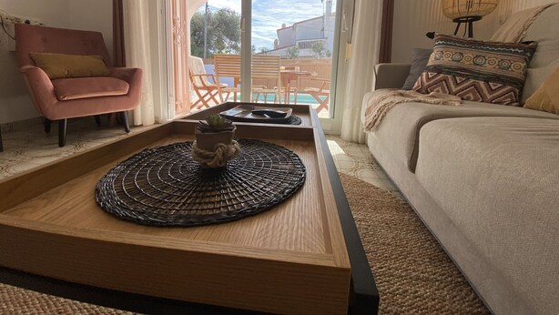 Uma sala de estar com sofá, uma cadeira, uma mesa de centro com uma planta e vista para o exterior através de uma porta de correr.