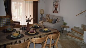 Uma sala de jantar com uma mesa de madeira posta para quatro pessoas, rodeada por cadeiras de madeira. Um sofá e uma pequena mesa lateral com uma planta são visíveis ao fundo.