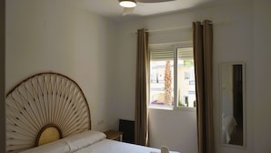 Quarto de hotel com uma cama, um ventilador de teto, uma janela com cortinas e vista para prédios.