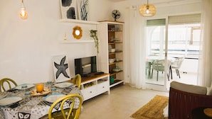 Uma área de refeições com uma mesa posta para quatro pessoas, uma televisão e uma estante com objetos decorativos.
