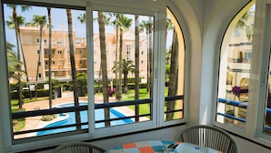 Quarto de hotel com vista para uma piscina, palmeiras e um edifício ao fundo.