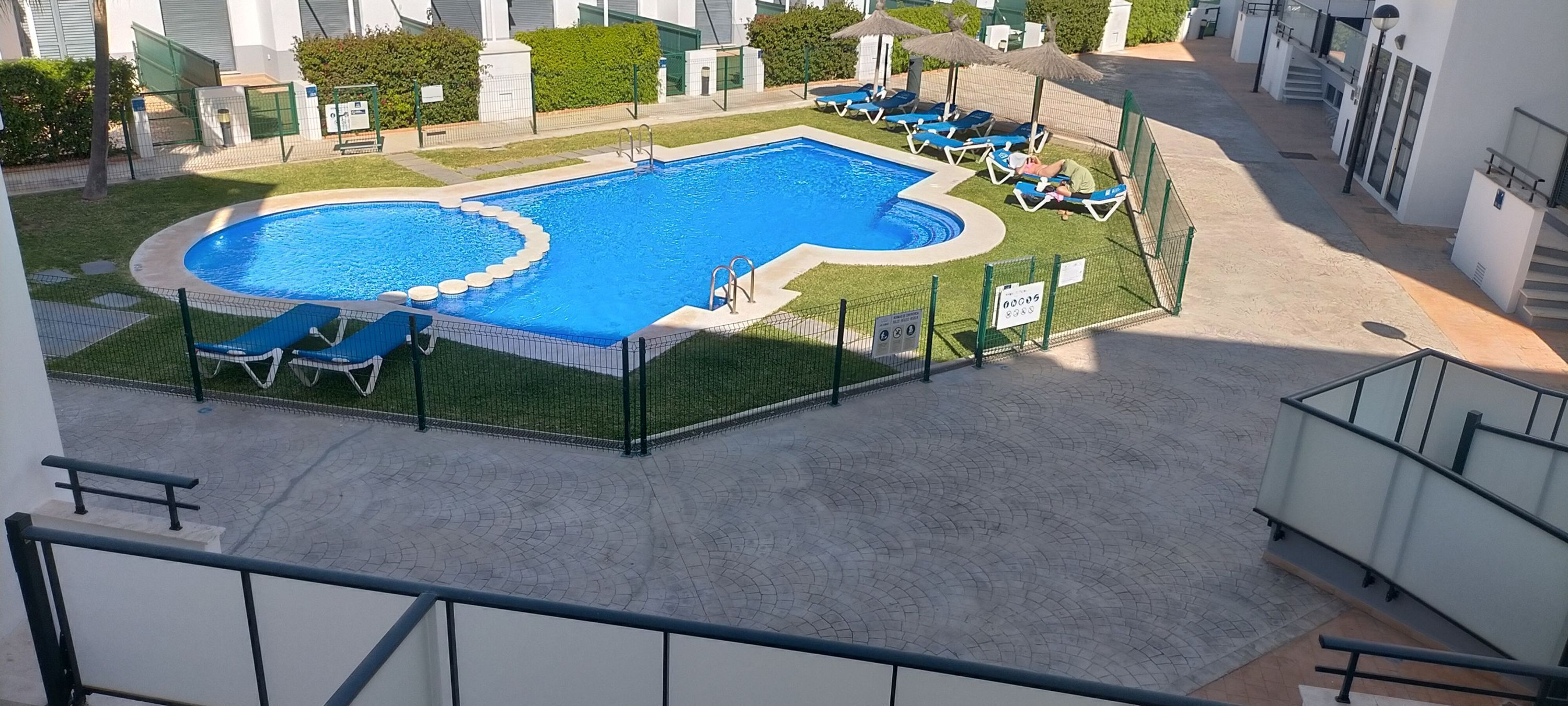 Piscina com espreguiçadeiras e área cercada, rodeada por um calçadão e vegetação.