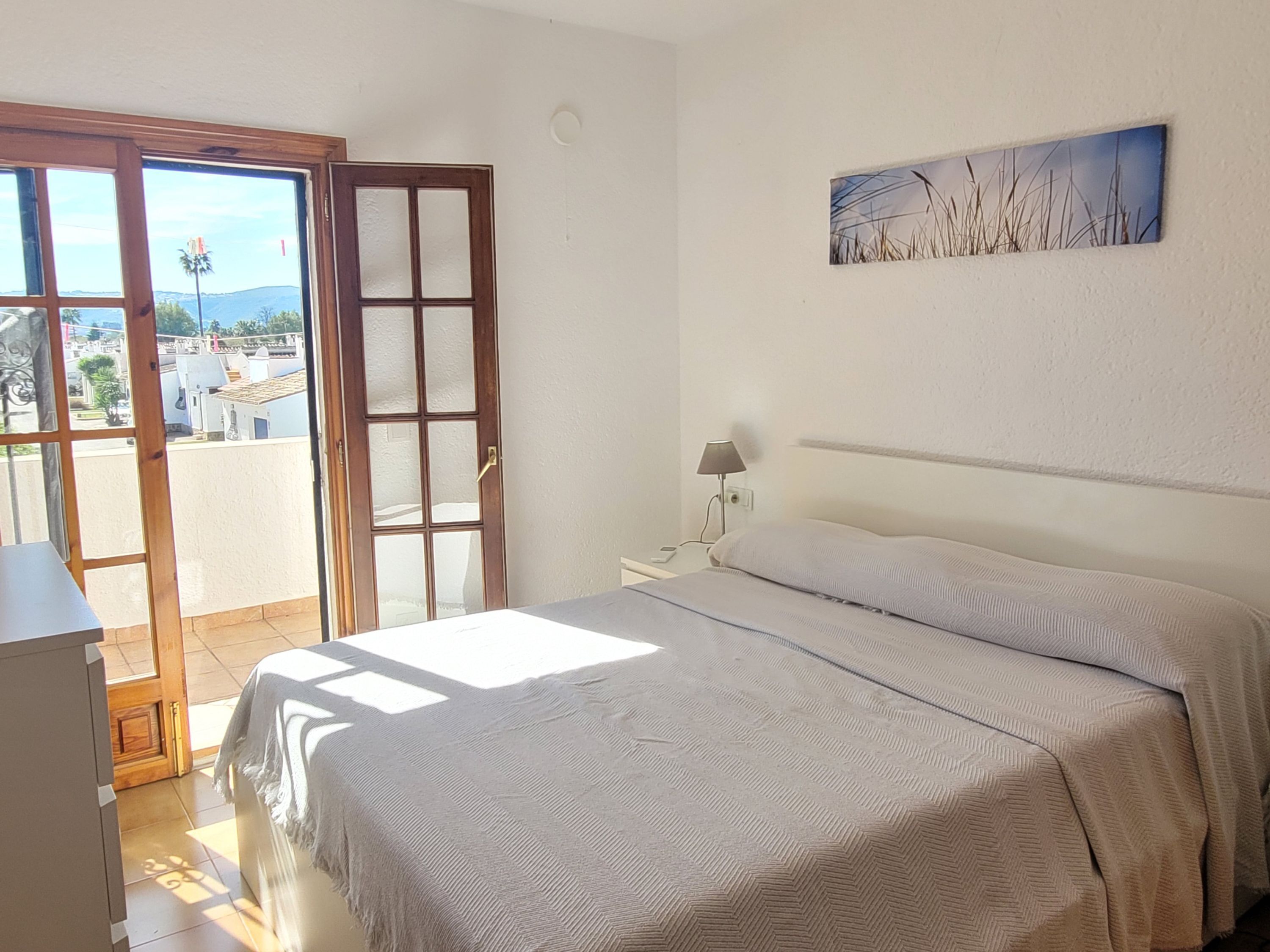 Quarto com cama, mesa de cabeceira, luminária, pintura e porta de vidro deslizante para uma varanda externa.