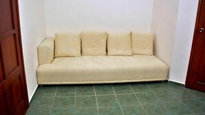 Um sofá bege com quatro almofadas num canto de um quarto com chão de azulejos verdes e paredes brancas.