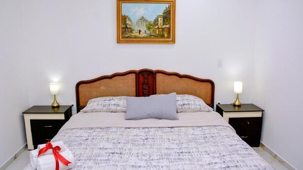 Uma cama bem arrumada com colcha estampada, duas mesas de cabeceira com luminárias, um quadro na parede e uma toalha enrolada com uma fita vermelha no chão.