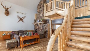 Interior de uma cabana de toras com lareira de pedra, escada de madeira, cabeça de veado montada e estante de livros.