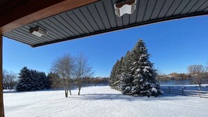Paisagem coberta de neve com céu claro, uma grande árvore perene e um corpo d'água ao fundo.