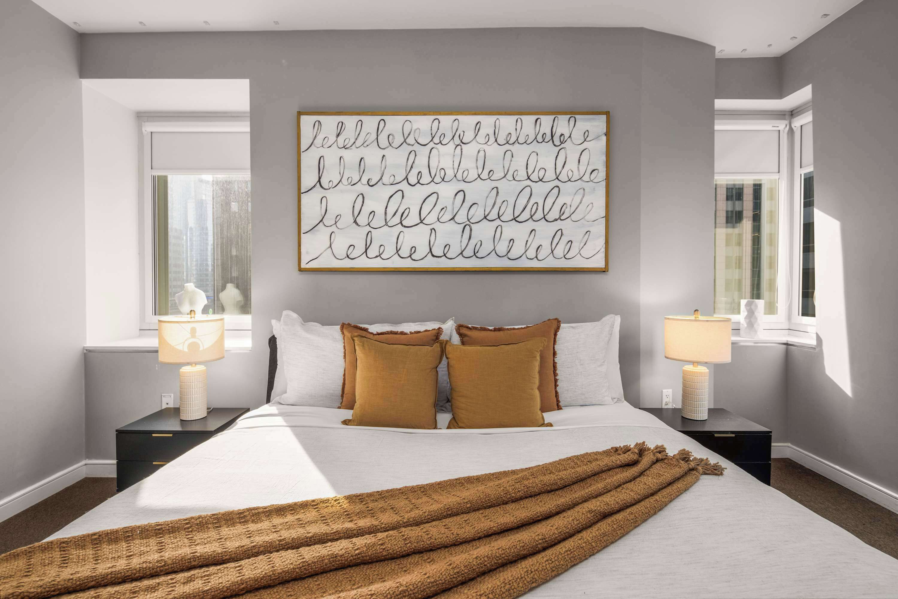 Quarto moderno com uma cama grande, duas mesas de cabeceira com abajures, um detalhe decorativo na parede e janelas amplas.