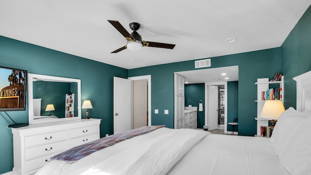 Quarto com paredes verde-água, uma cama branca, um ventilador de teto, cômoda branca e uma estante de livros.