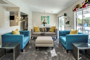 Uma sala de estar moderna com dois sofás azuis, um sofá cinza e um banquinho bege. Há um candelabro, um quadro e uma planta na parede.