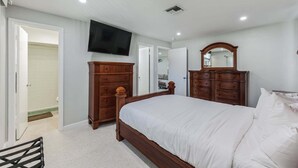 Quarto com cama, cómoda de madeira, televisão montada na parede e espelho sobre a cómoda.