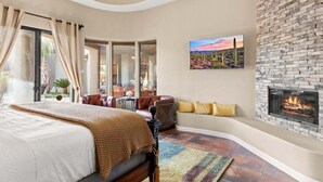 Quarto com uma cama grande, lareira, vista para o exterior e área de refeições visível através de portas de vidro.