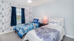 Quarto com duas camas, janela com cortinas azuis, parede com padrão de planta baixa e mesa de cabeceira com luminária.