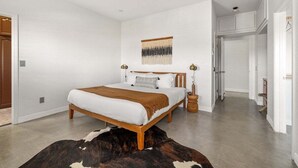 Quarto com uma cama grande, cabeceira de madeira, tapete de couro e luminária fixada na parede, além de um corredor que dá para outro cômodo.