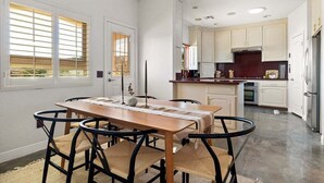 Uma área de refeições com mesa e cadeiras de madeira, uma cozinha com armários brancos e eletrodomésticos de aço inoxidável, e uma janela com persianas.