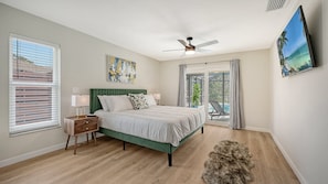 Quarto com cama, mesas de cabeceira, ventoinha no teto, um quadro e vista para uma piscina através de uma porta de vidro deslizante.