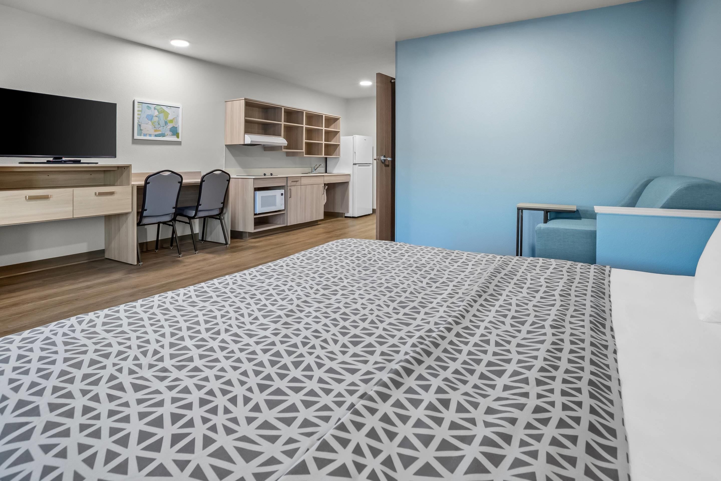 Quarto moderno com cama, escrivaninha, cadeira, televisão, área de cozinha e parede azul.