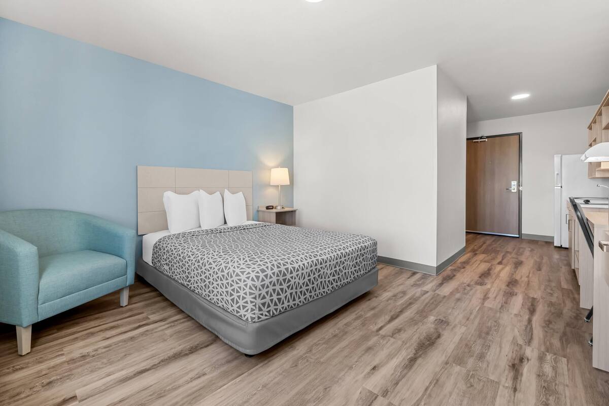 Quarto de hotel moderno com cama, sofá, escrivaninha e cozinha compacta.