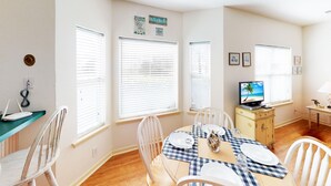 Uma sala de jantar com uma mesa posta para quatro pessoas, cadeiras brancas, uma televisão em um móvel e uma janela com persianas.