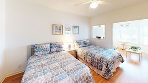 Quarto com duas camas, ventilador no teto, uma mesinha com uma planta e janela com persianas.