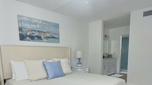 Quarto com cama, mesa de cabeceira, luminária, pintura e banheiro visível por uma porta aberta.