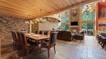Uma sala de jantar ampla com mesa e cadeiras de madeira, parede de pedra e uma grande janela com vista para uma floresta.