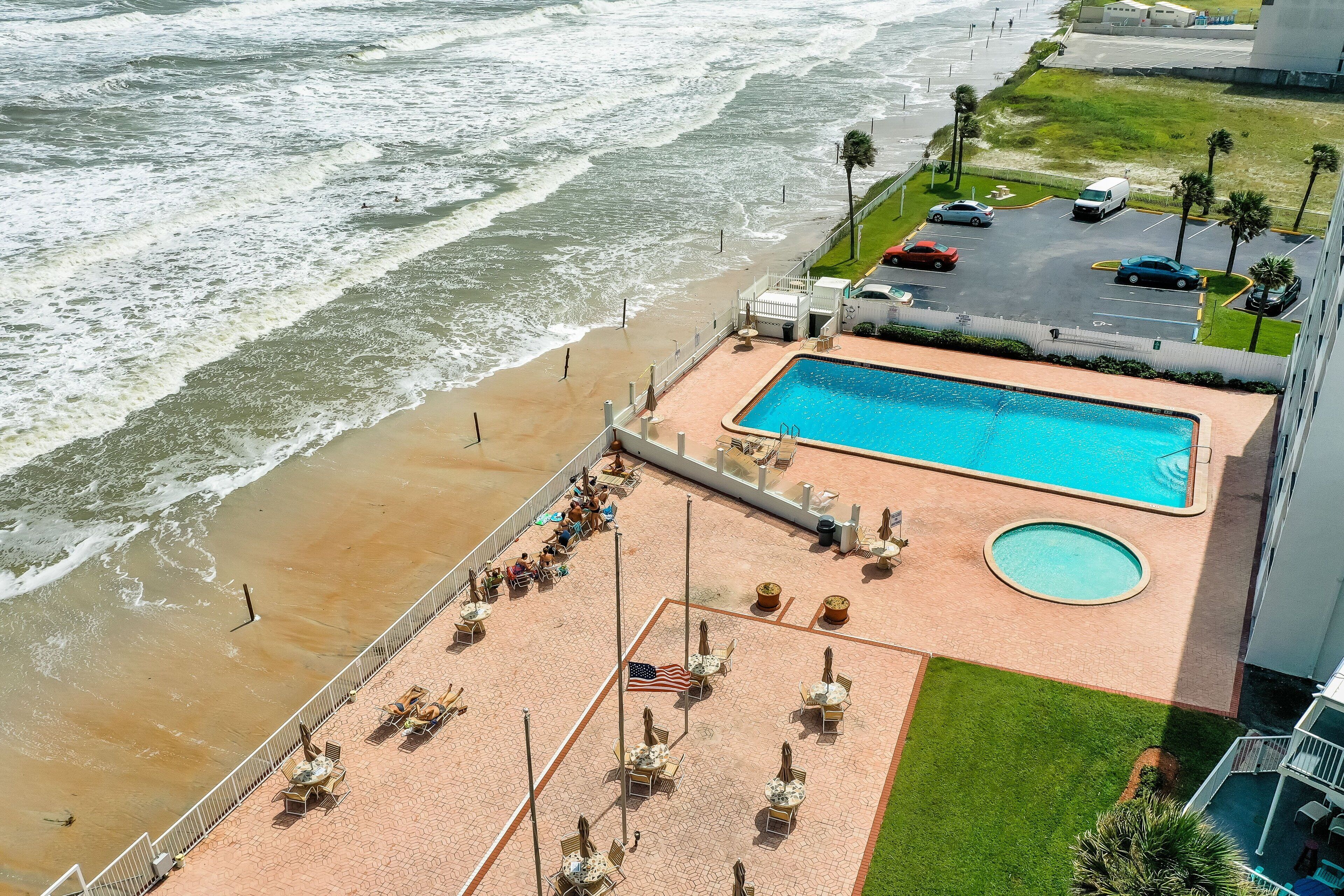 Vista aérea de um hotel na praia com piscina, área de estar à beira-mar e estacionamento.
