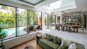 Uma sala de estar ampla com uma grande janela com vista para uma piscina, uma área de jantar com uma mesa comprida e uma cozinha visível através de portas de vidro.