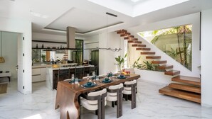 Uma cozinha moderna com ilha central, um espaço de jantar com uma mesa de madeira comprida e uma escada com guarda-corrimão de vidro que dá para um andar superior.