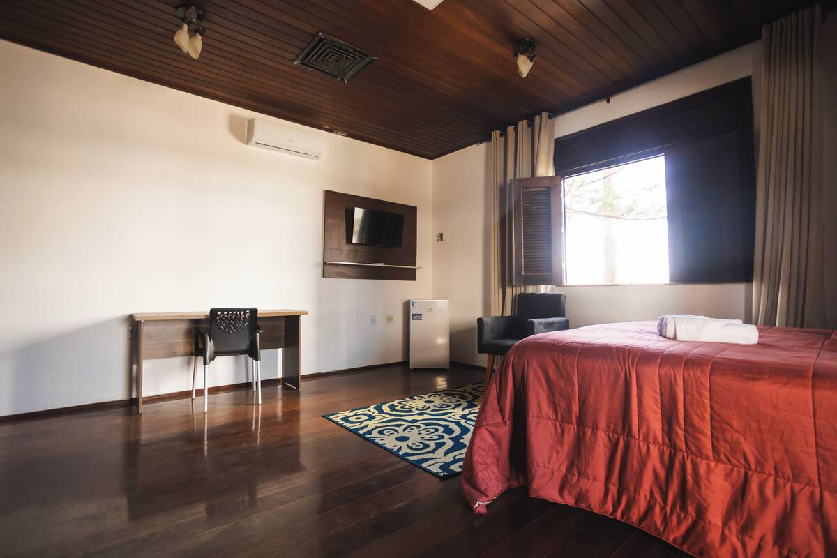 Um quarto com cama, escrivaninha, cadeira, televisão e janela com cortinas.