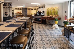 Restaurante com uma mesa comunitária grande, cadeiras, área de bar e plantas decorativas.