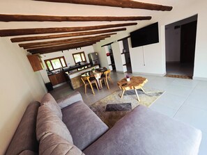 Uma sala moderna com sofá, área de jantar com cadeiras, cozinha ao fundo e televisão de tela plana na parede.