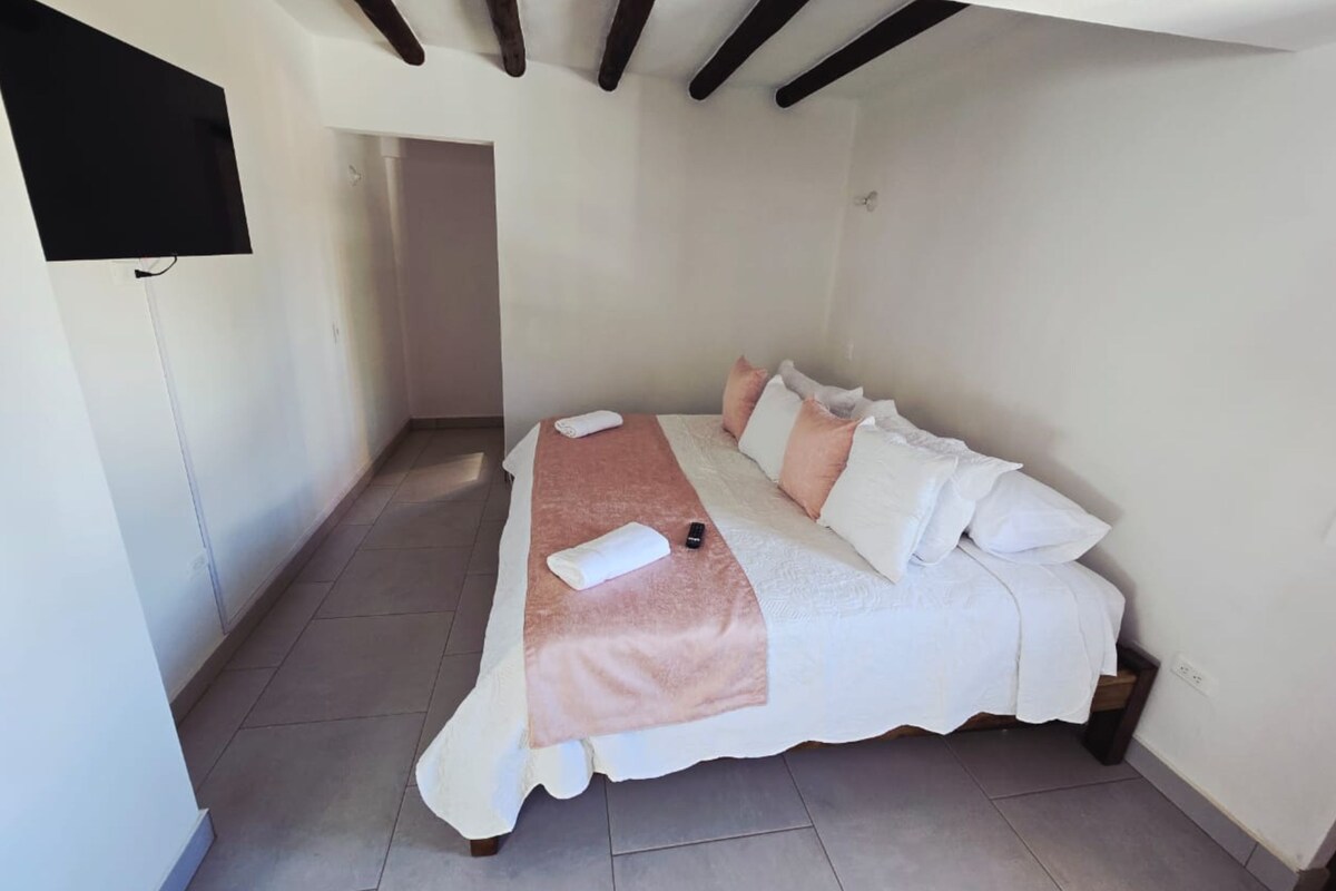 Quarto com cama, almofadas, cobertor, mesa pequena e televisão na parede.