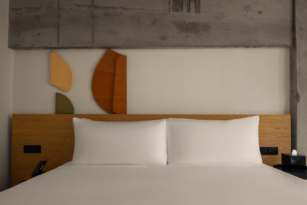 Uma cama com cabeceira de madeira, duas almofadas brancas e uma mesa de cabeceira com um telefone e um abajur.