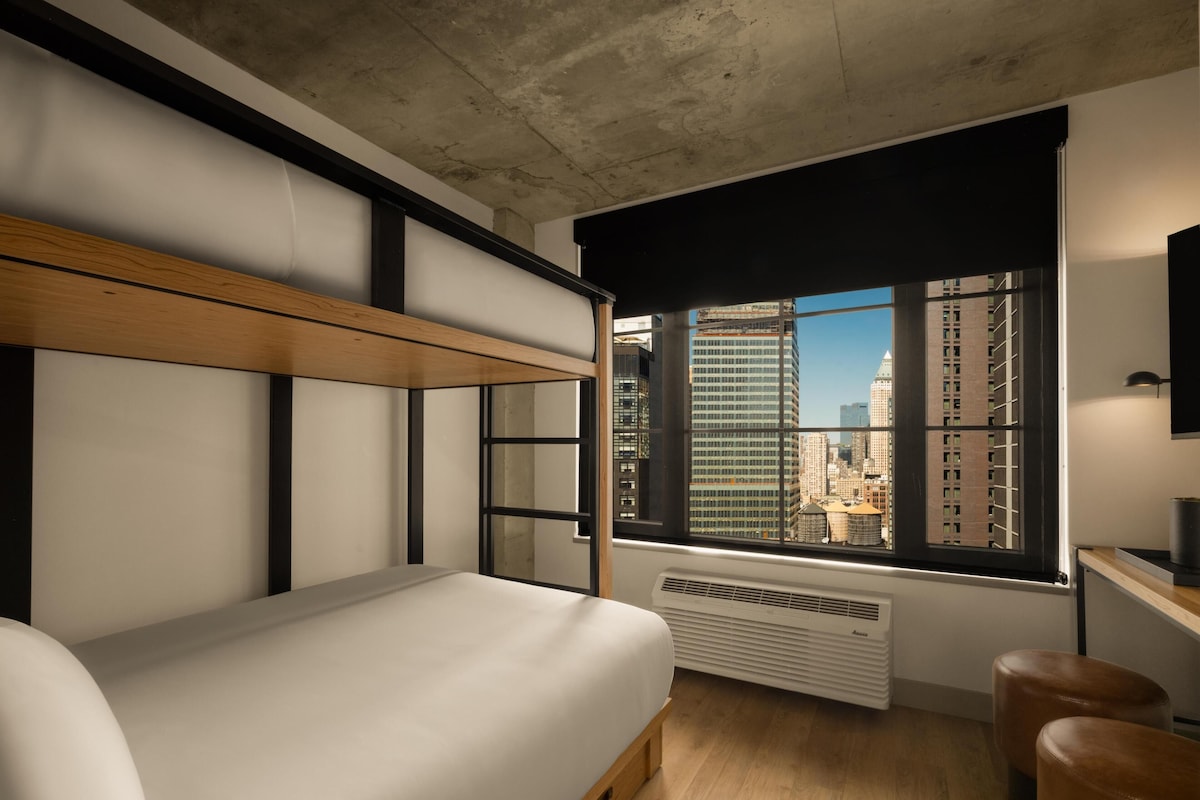 Quarto moderno com cama beliche, escrivaninha, cadeira e uma grande janela com vista para a cidade.