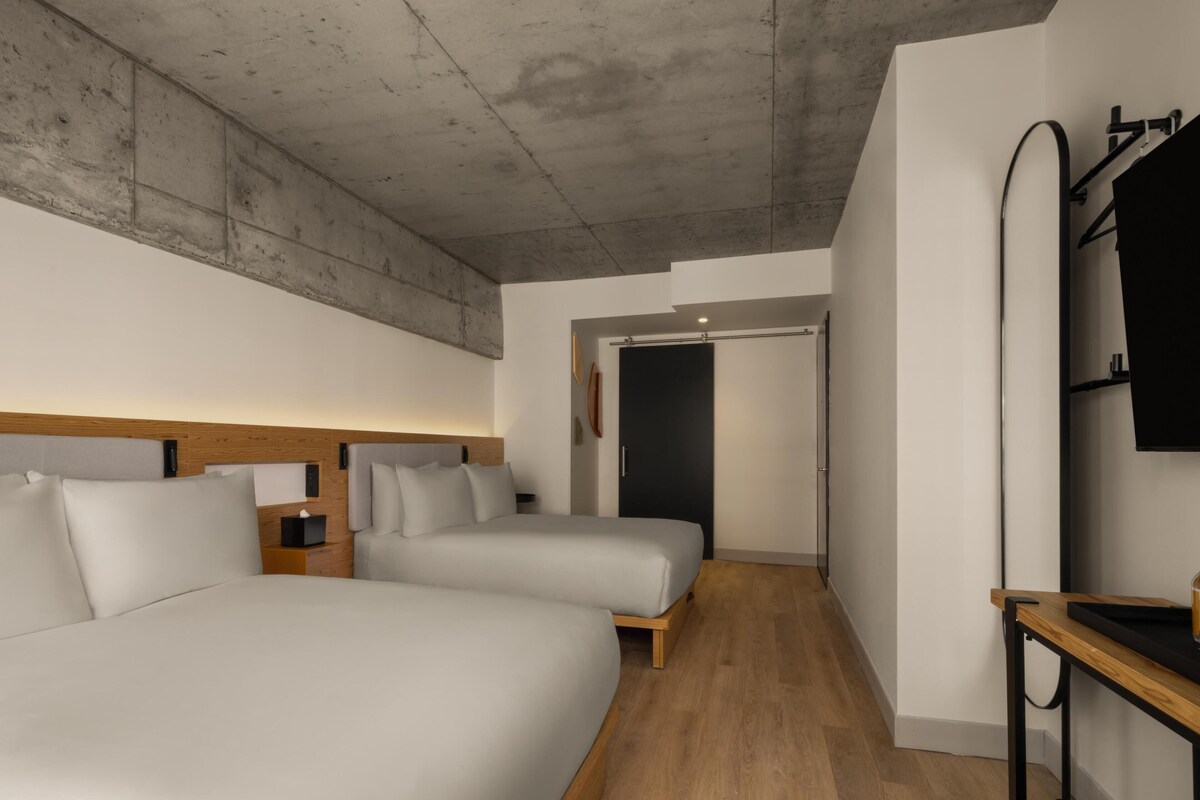 Quarto de hotel com duas camas, uma escrivaninha de madeira, televisão de tela plana e teto de concreto.
